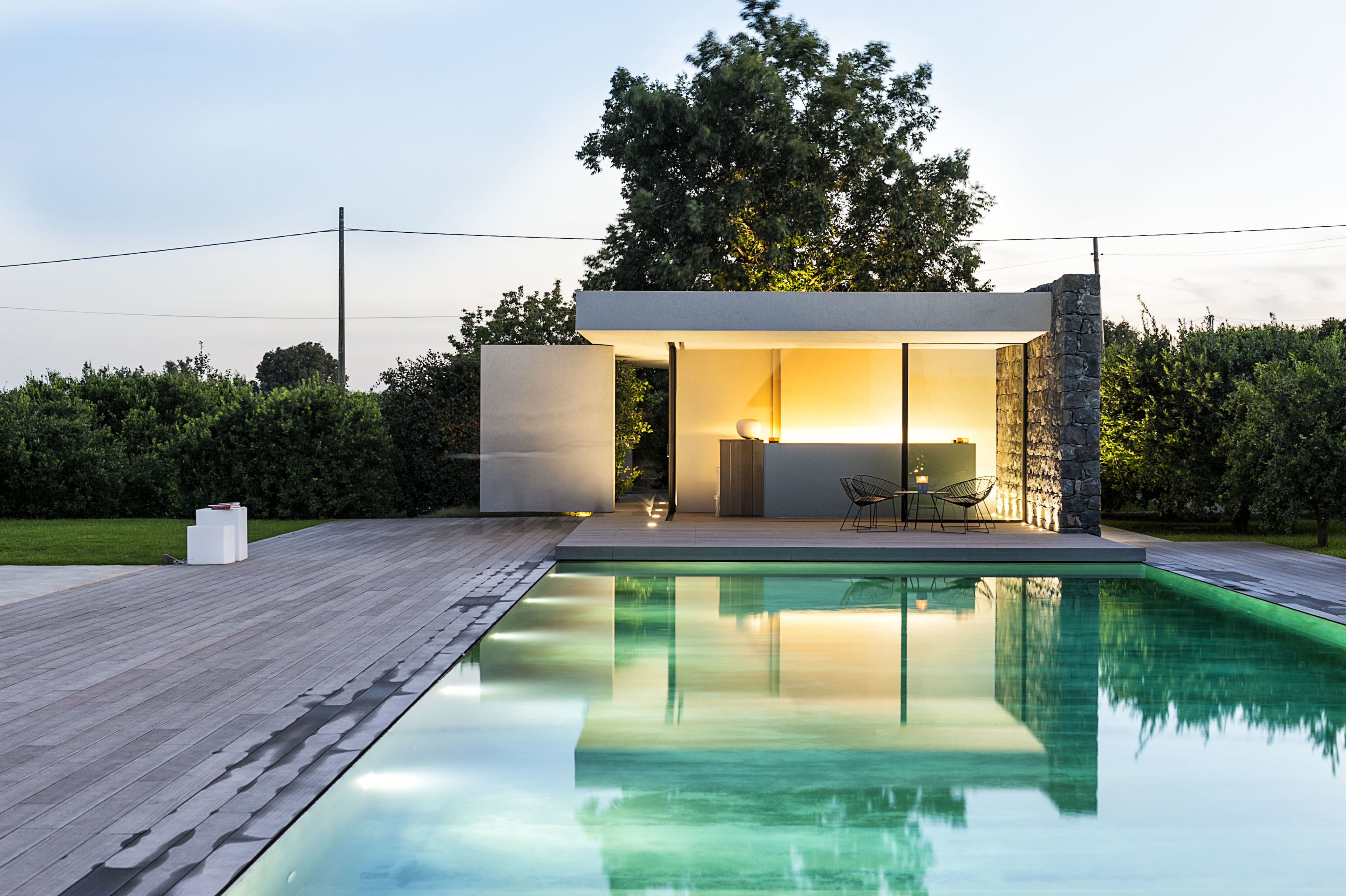 Uma piscina moderna ao ar livre, com um bangalô minimalista, deck de madeira e uma parede de pedra.