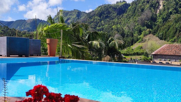 Piscina azul límpida com deck de madeira, rodeada por vegetação exuberante e montanhas ao fundo.