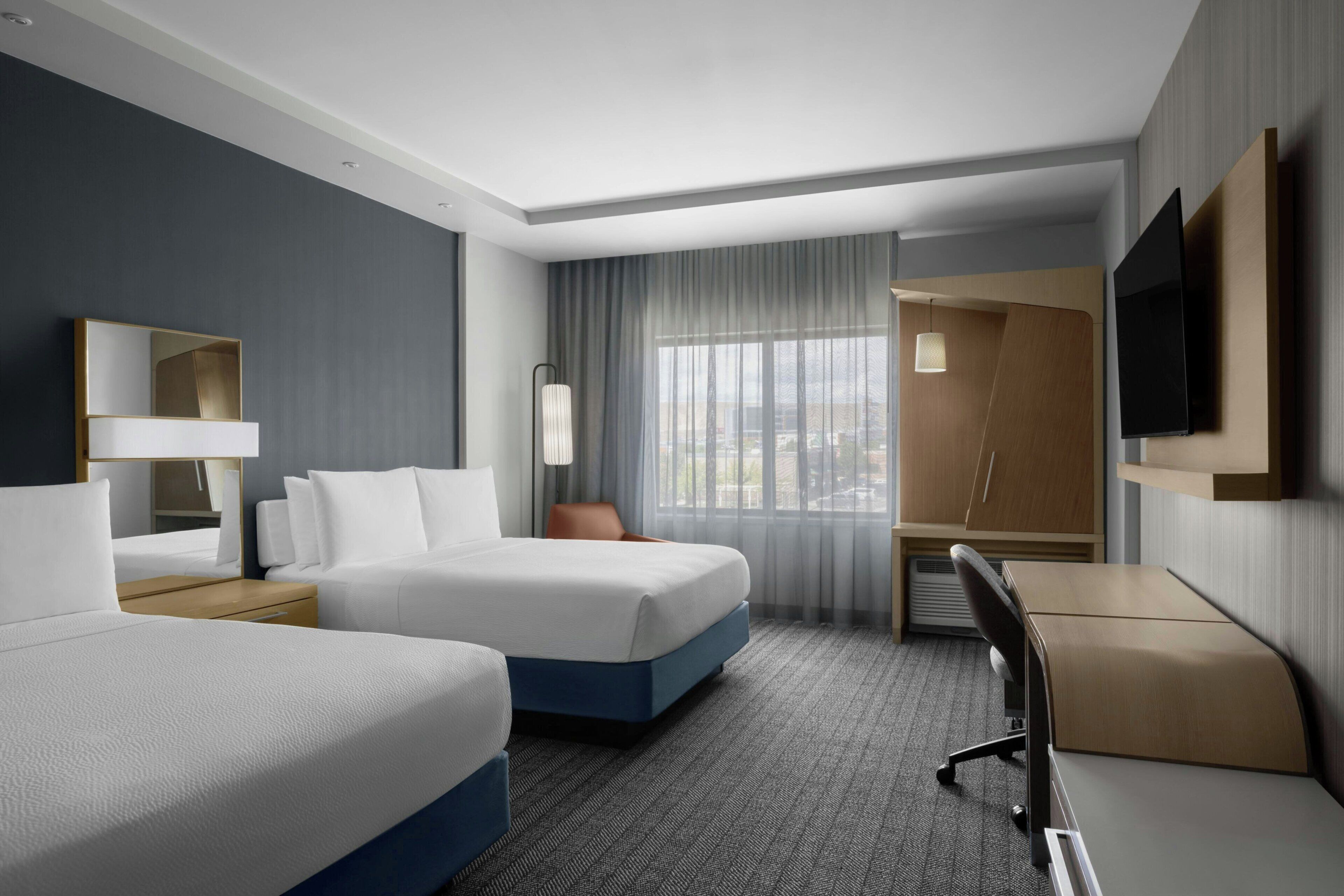 Quarto de hotel com duas camas, uma escrivaninha, uma cadeira, televisão e janela com cortinas.