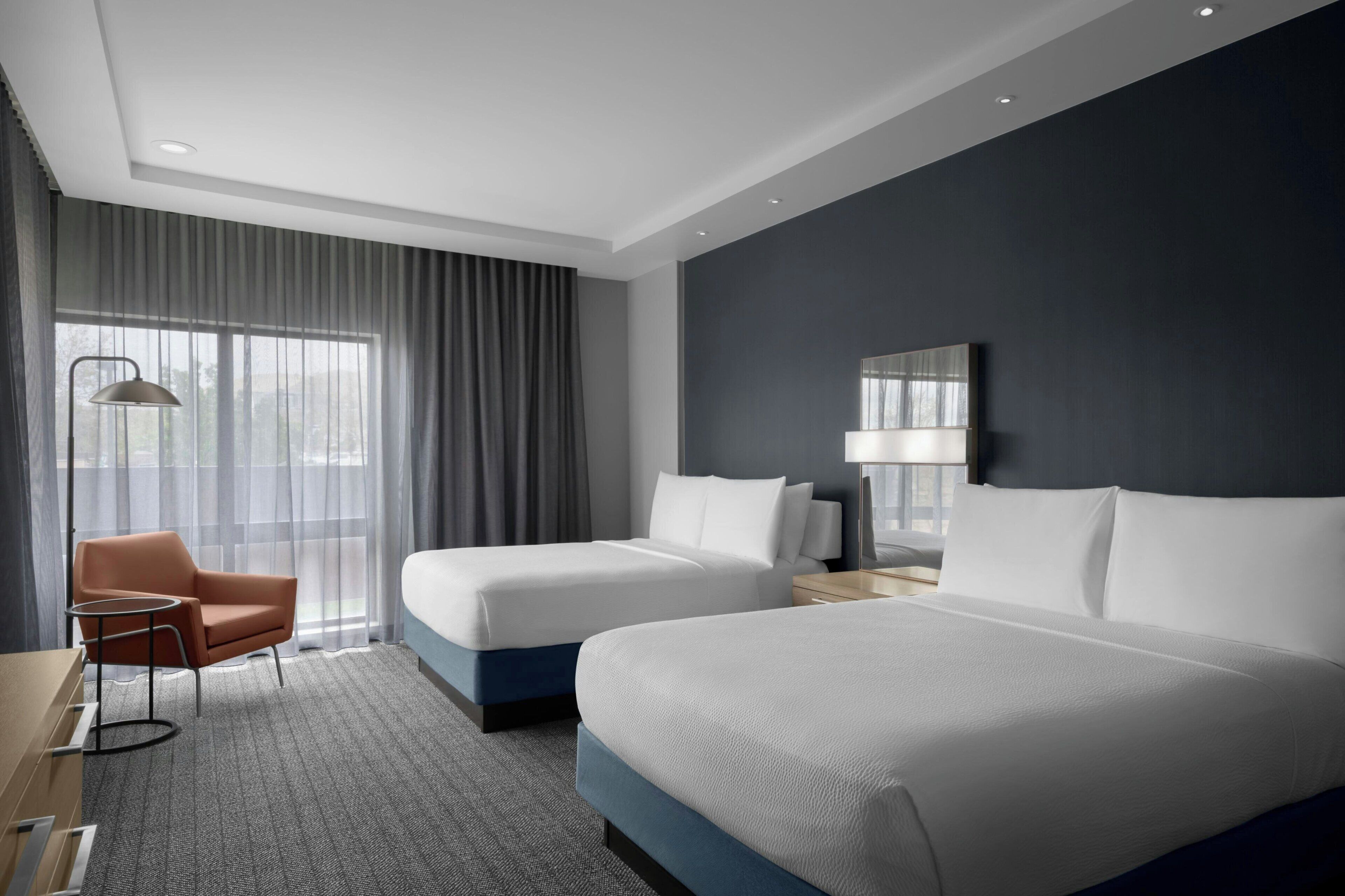 Quarto de hotel moderno com uma cama grande, uma cama menor, uma escrivaninha, uma cadeira e uma janela ampla com cortinas.