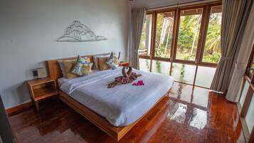 Cama grande com estrutura de madeira, roupa de cama branca e almofadas decorativas. Tem uma mesa de cabeceira de madeira com uma luminária e uma obra de arte na parede. O quarto tem uma janela grande com cortinas e vista para o verde lá fora.