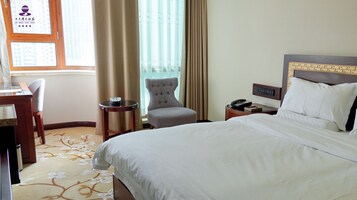 Quarto de hotel com cama, poltrona, escrivaninha, mesa de cabeceira e janela com cortinas.