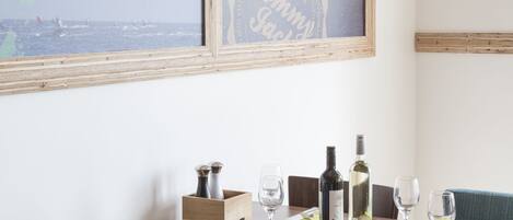 Uma área de jantar com mesa de madeira posta para dois, cadeiras estofadas em azul e obras de arte emolduradas retratando pipas e uma cena de praia.