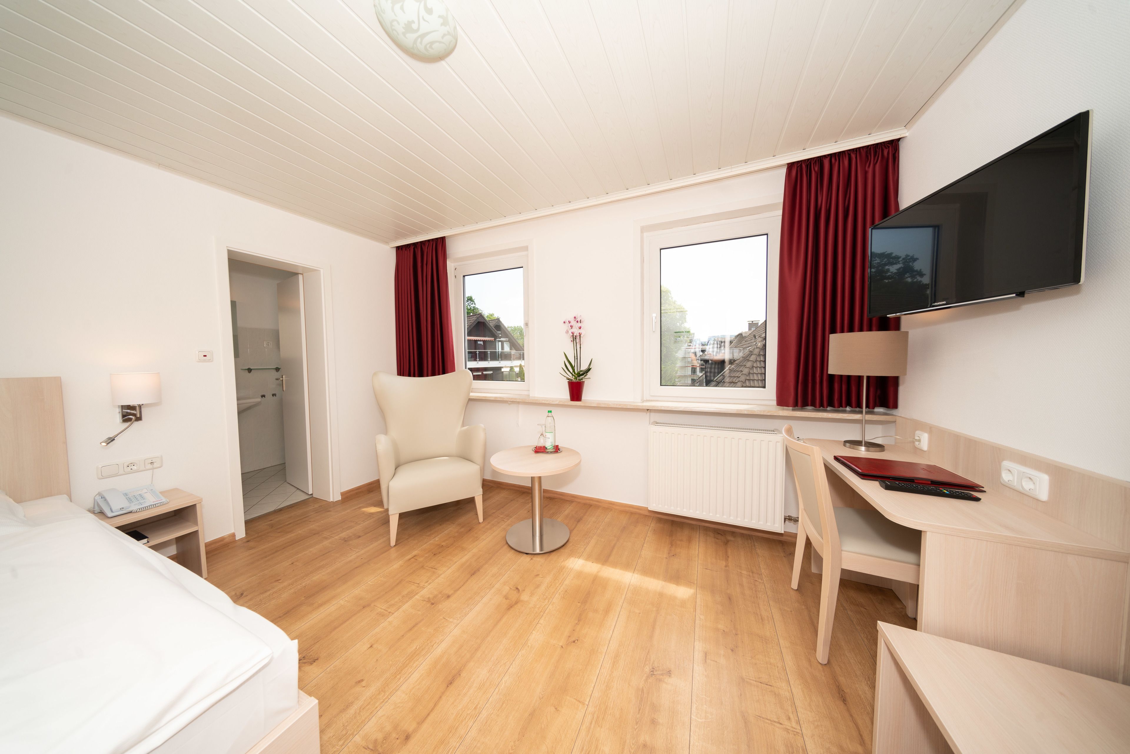 Quarto de hotel com cama, escrivaninha, cadeira, televisão e janela com cortinas.