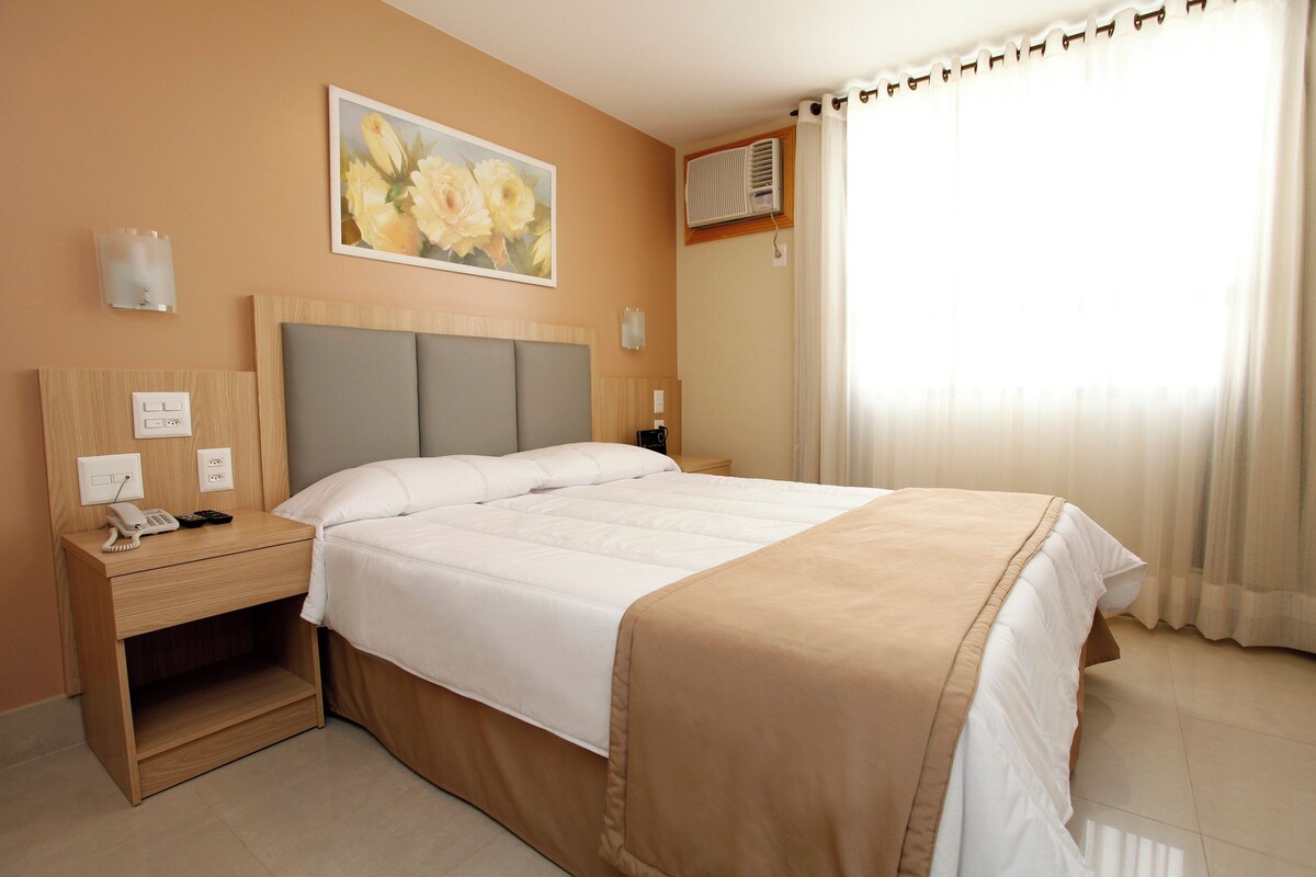 Quarto de hotel com cama, mesas de cabeceira, um quadro, ar condicionado e janela com cortinas.