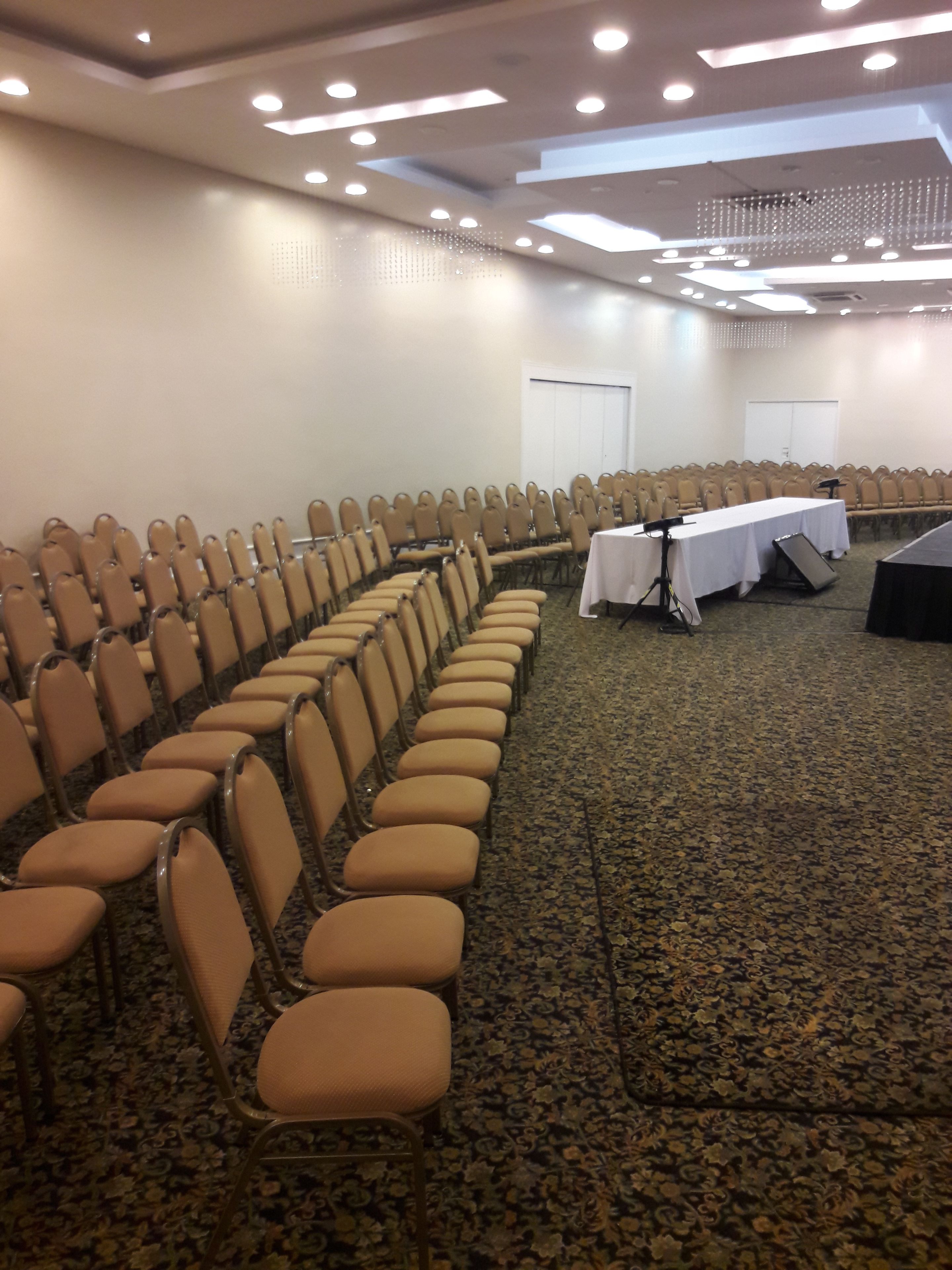 Uma sala de conferência com fileiras de cadeiras bege, uma mesa branca com um laptop e piso carpeteado.