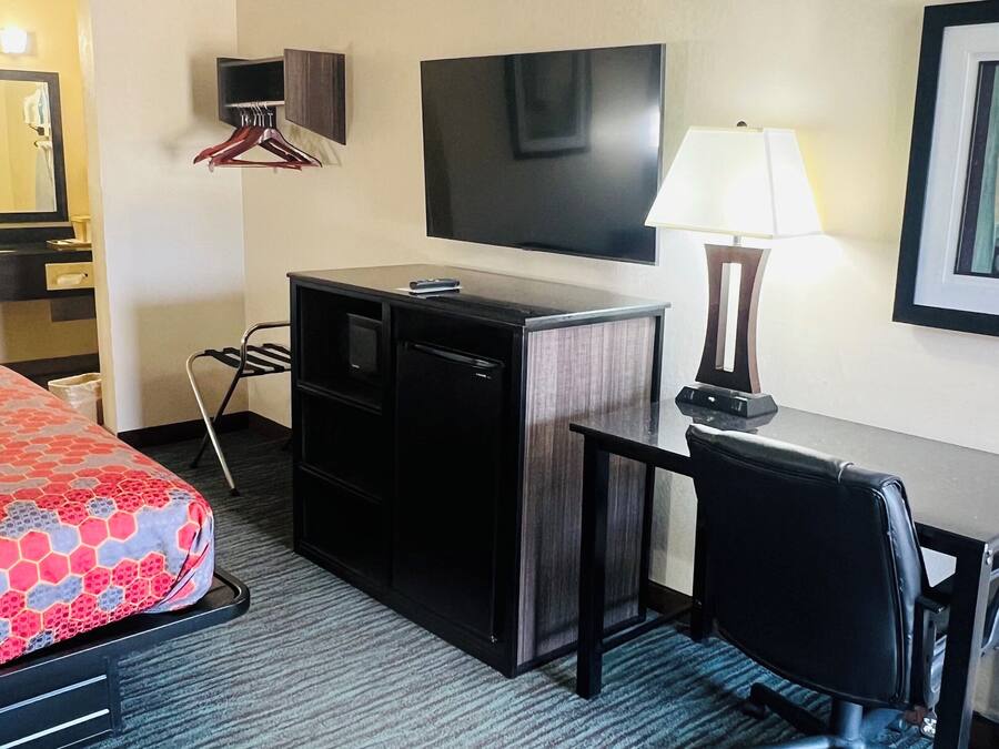 Quarto de hotel com cama, escrivaninha, cadeira, televisão, abajur e cabideiro.