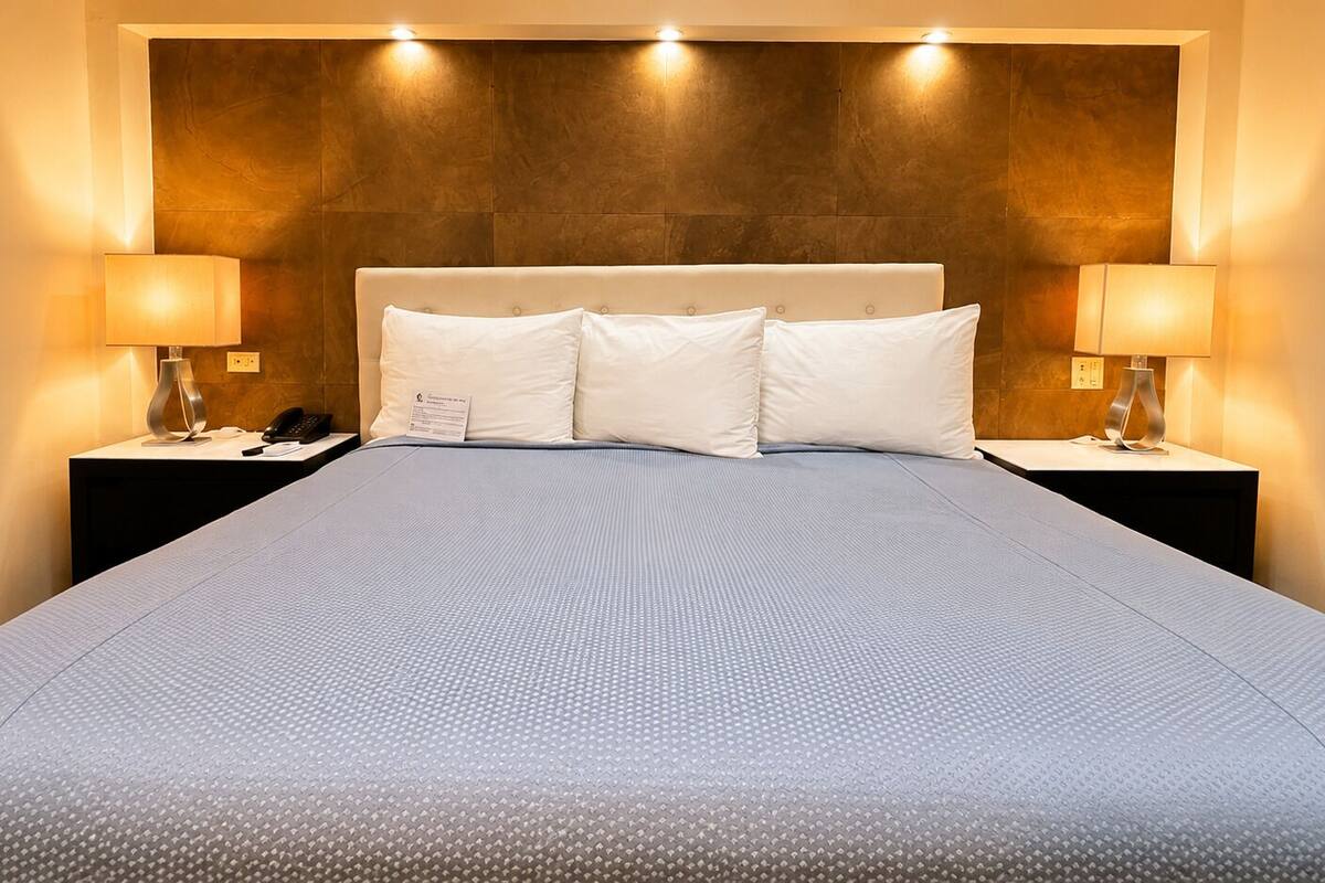Quarto de hotel com uma cama grande, duas mesas de cabeceira, uma luminária fixada na parede e um telefone.