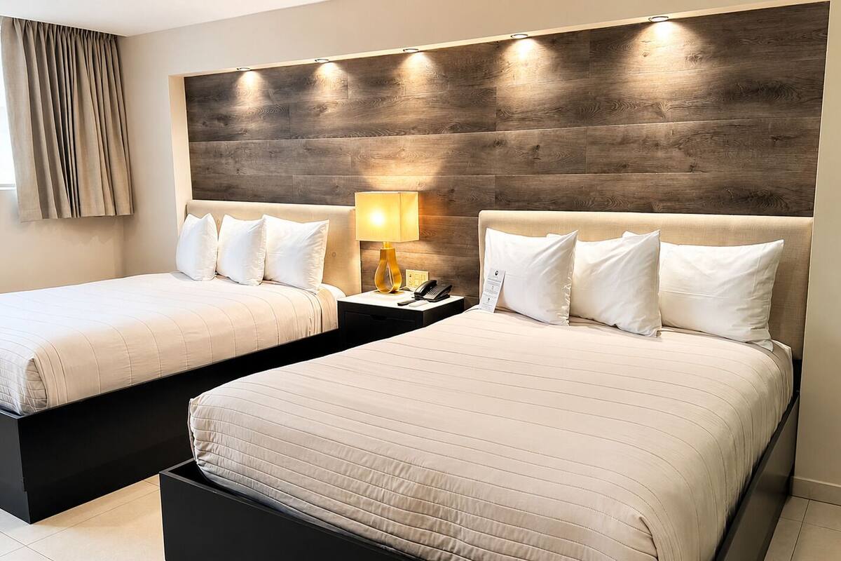 Quarto de hotel com duas camas, cabeceira de madeira, luminárias de apoio e uma janela com cortinas.