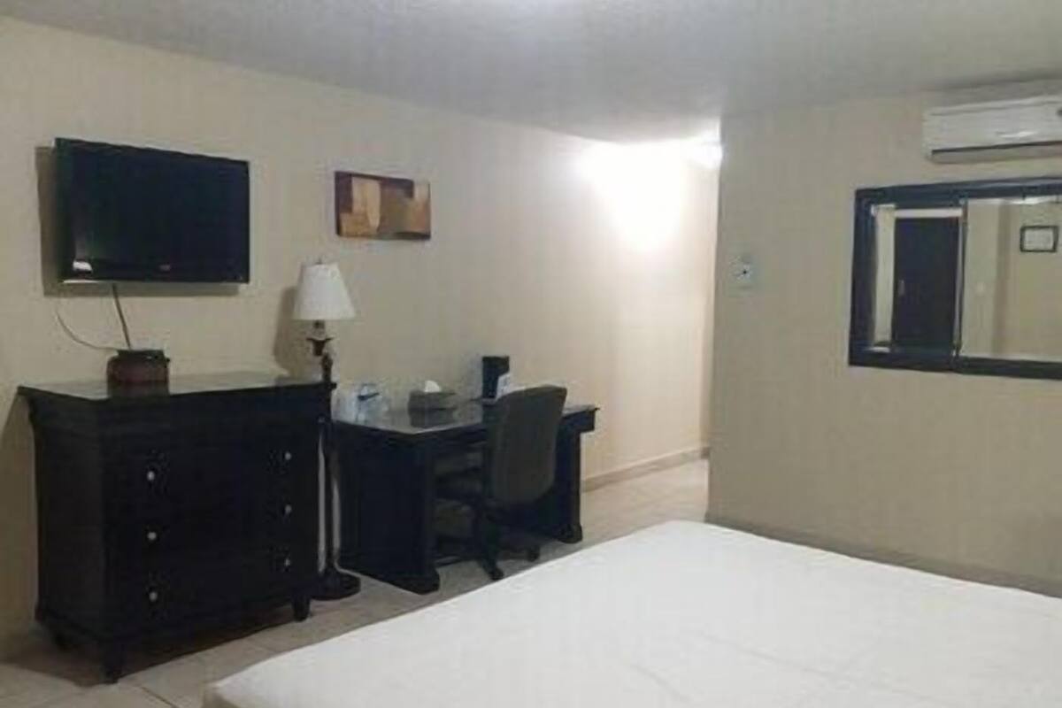 Quarto de hotel com cama, escrivaninha, cadeira, televisão e luminária.