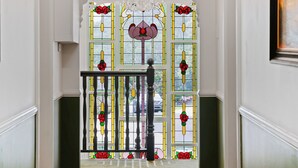 Um corredor com carpete estampado, um candelabro e uma porta com vitrais de motivos florais.