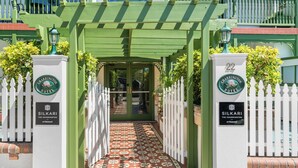 Entrada de hotel com pérgola verde, cerca branca de piquete e um caminho de ladrilho que leva à porta.