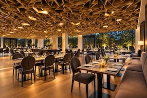 Uma sala de jantar espaçosa, com pé-direito alto e iluminação moderna. Mesas postas para refeições, com cadeiras.