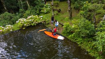 Uma pessoa praticando paddleboarding em um rio, cercada por vegetação exuberante e arbustos de hortênsia.
