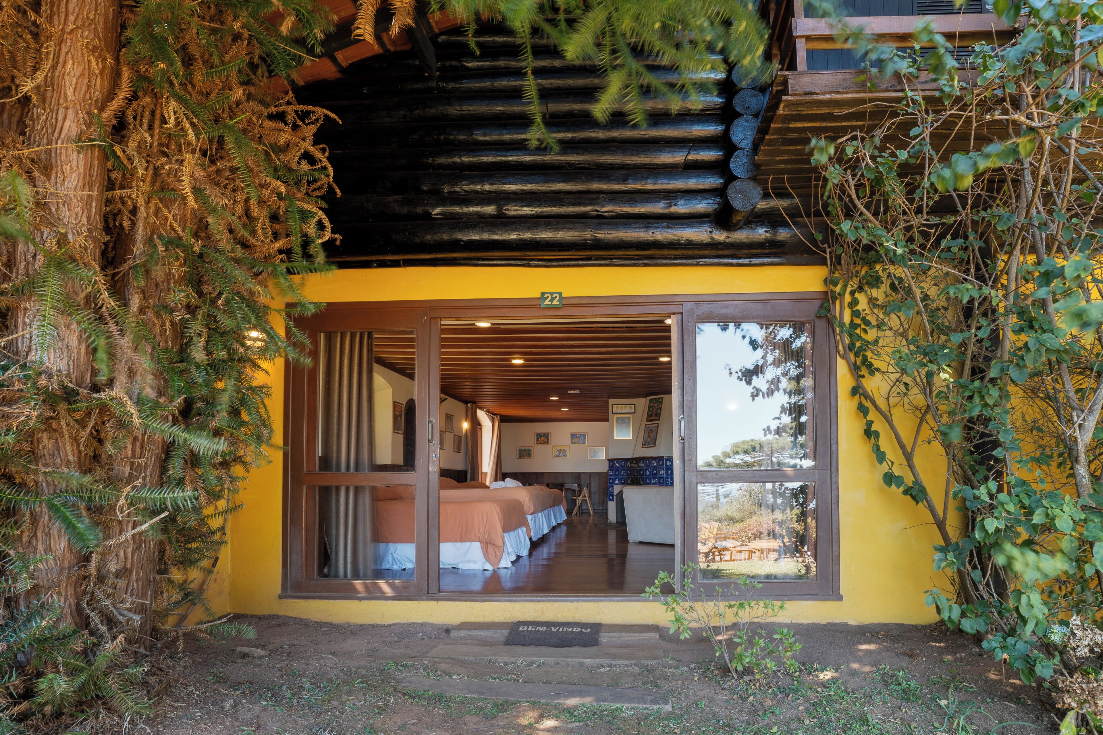 Entrada de uma pousada com fachada de madeira, porta de vidro que dá para um quarto com camas e área de refeições.