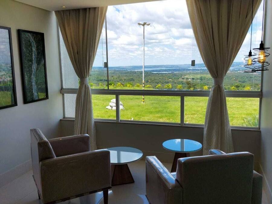 Quarto com duas poltronas e uma mesa redonda pequena, janelas grandes com cortinas e vista para uma paisagem verde.
