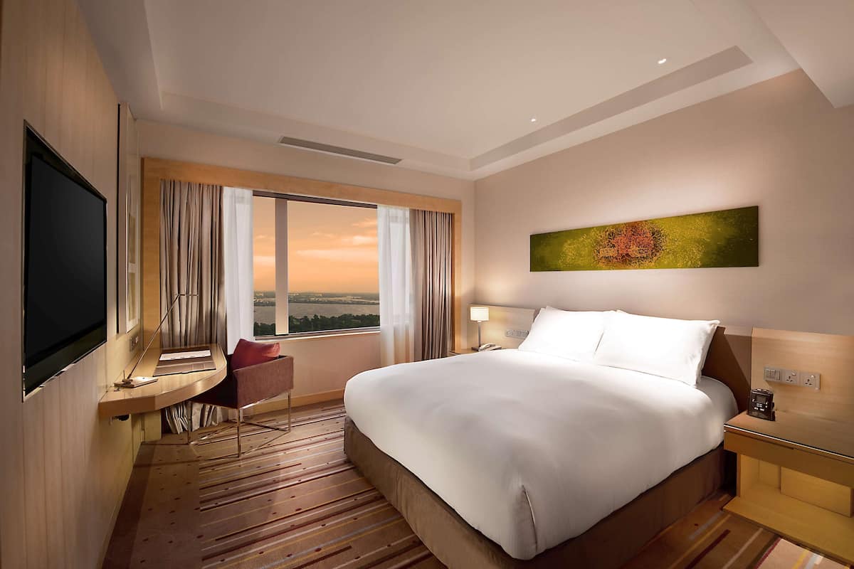 Quarto de hotel com uma cama grande, uma mesa com cadeira, televisão, janela com cortinas e um quadro na parede.