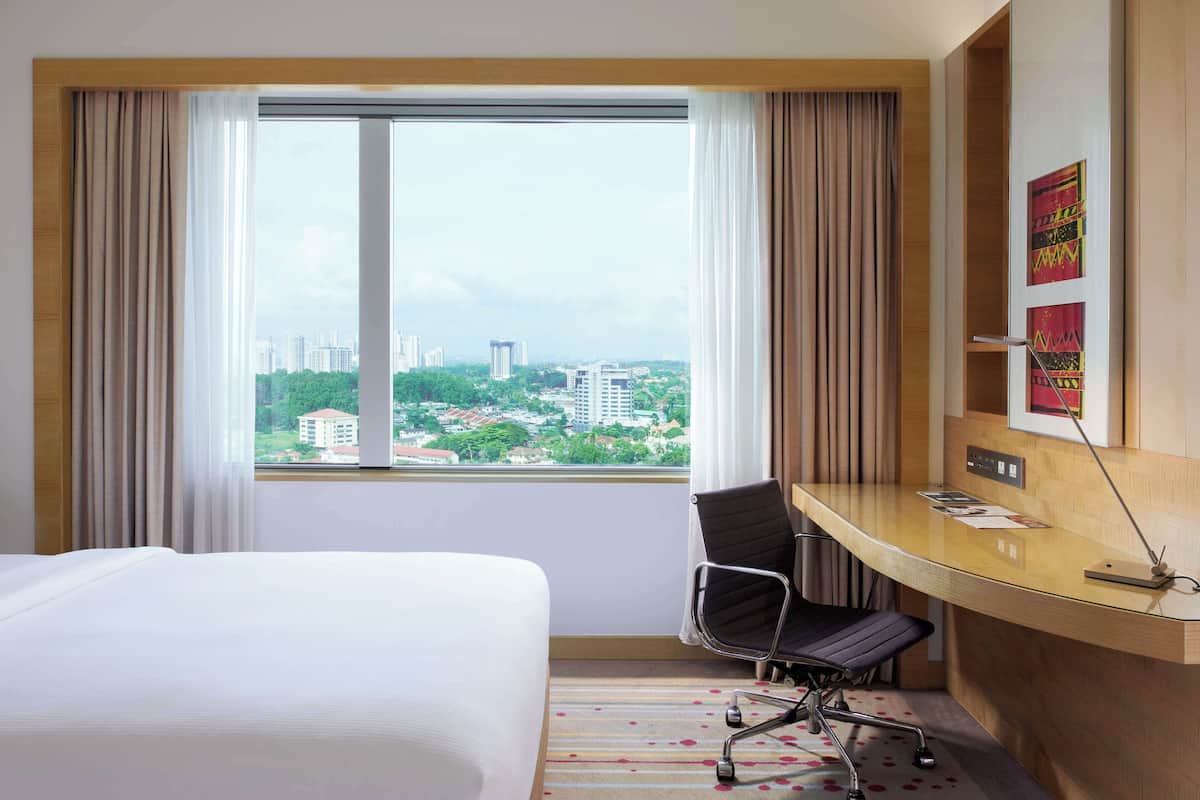 Quarto de hotel com cama, escrivaninha, cadeira, janela grande com cortinas e vista para a cidade.