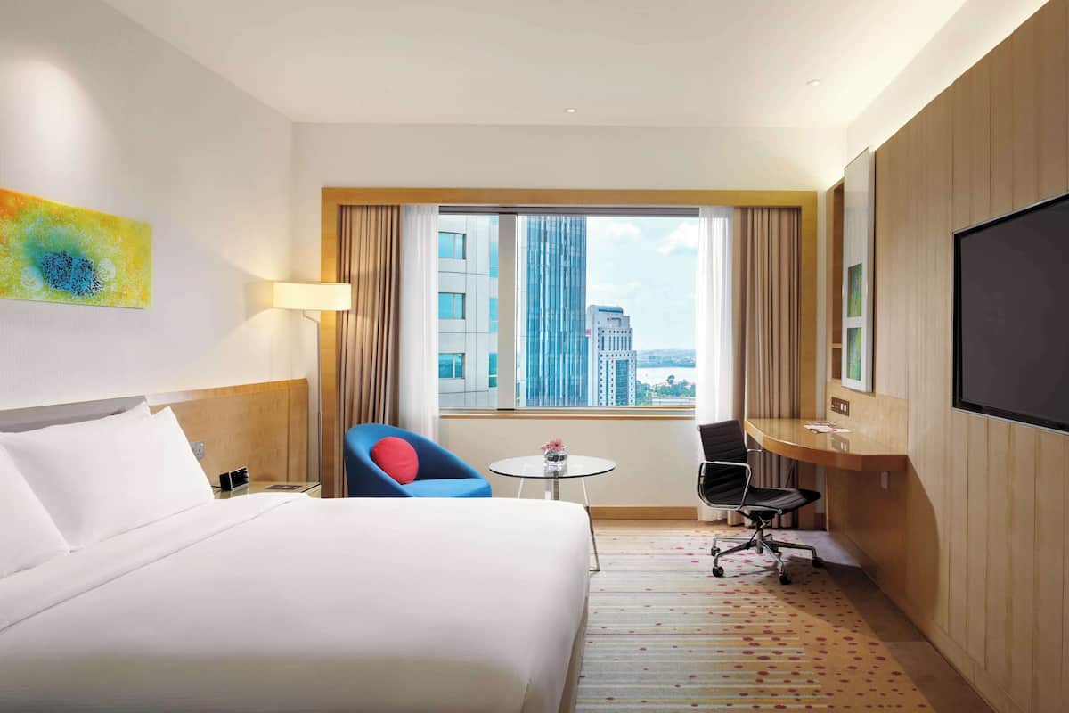 Quarto de hotel com cama, escrivaninha, cadeira, mesinha, televisão e janela com vista para a cidade.