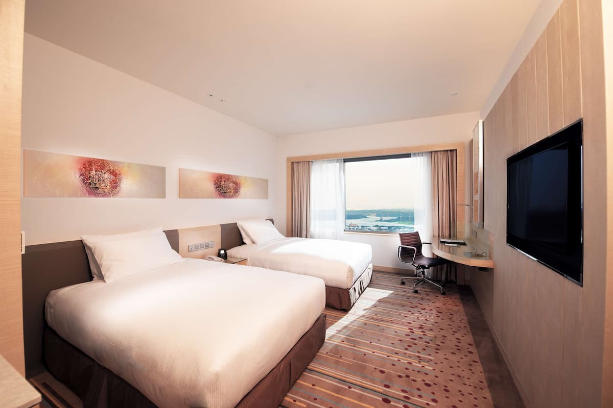 Quarto de hotel com duas camas, televisão, escrivaninha e uma grande janela com vista.