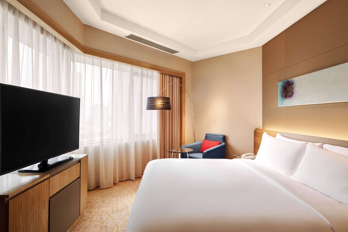 Quarto de hotel com uma cama grande, uma TV de tela plana sobre um móvel de madeira, uma cadeira com almofada vermelha, uma mesinha e um quadro na parede.