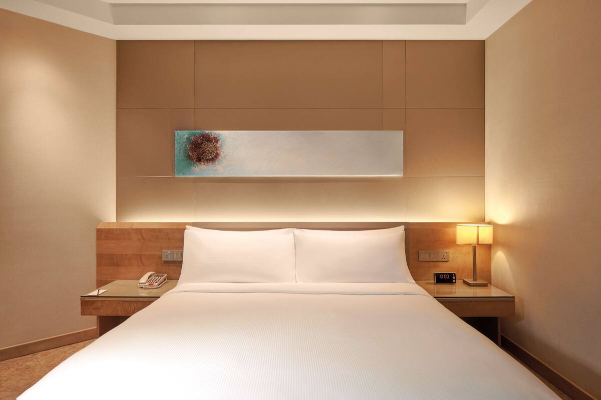 Quarto de hotel com cama grande, duas mesas de cabeceira, uma obra de arte pendurada na parede e um criado-mudo com luminária.