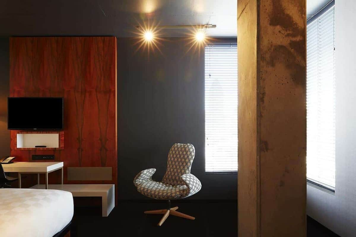 Quarto de hotel moderno com uma cama grande, uma televisão presa à parede, uma escrivaninha com cadeira e uma poltrona estampada.