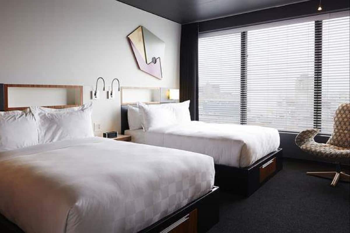 Quarto de hotel com duas camas, uma cadeira e uma janela grande com persianas.