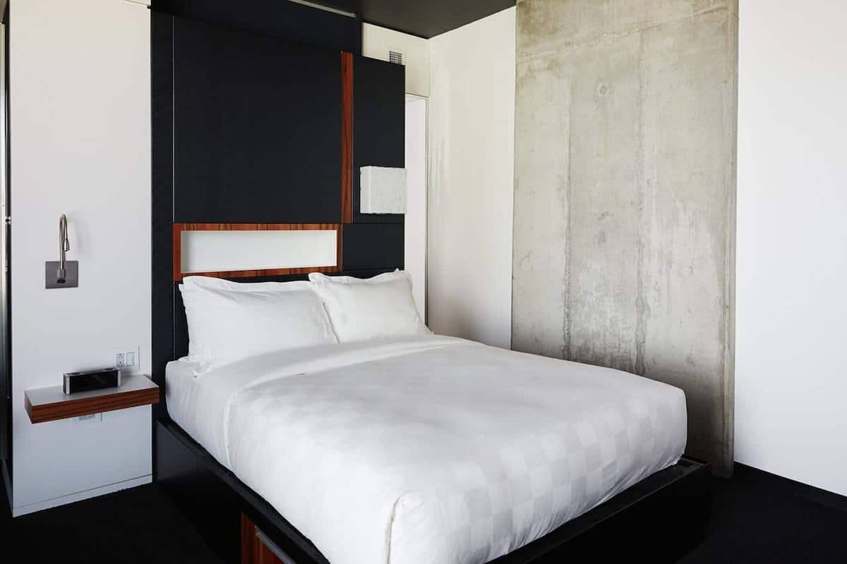 Quarto de hotel moderno com uma cama grande, cabeceira de madeira, mesa de cabeceira e uma parede de concreto como destaque.