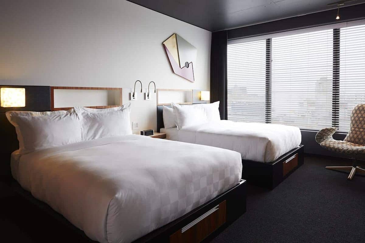Quarto de hotel com duas camas, uma cadeira moderna, uma luminária na parede e uma janela com persianas.