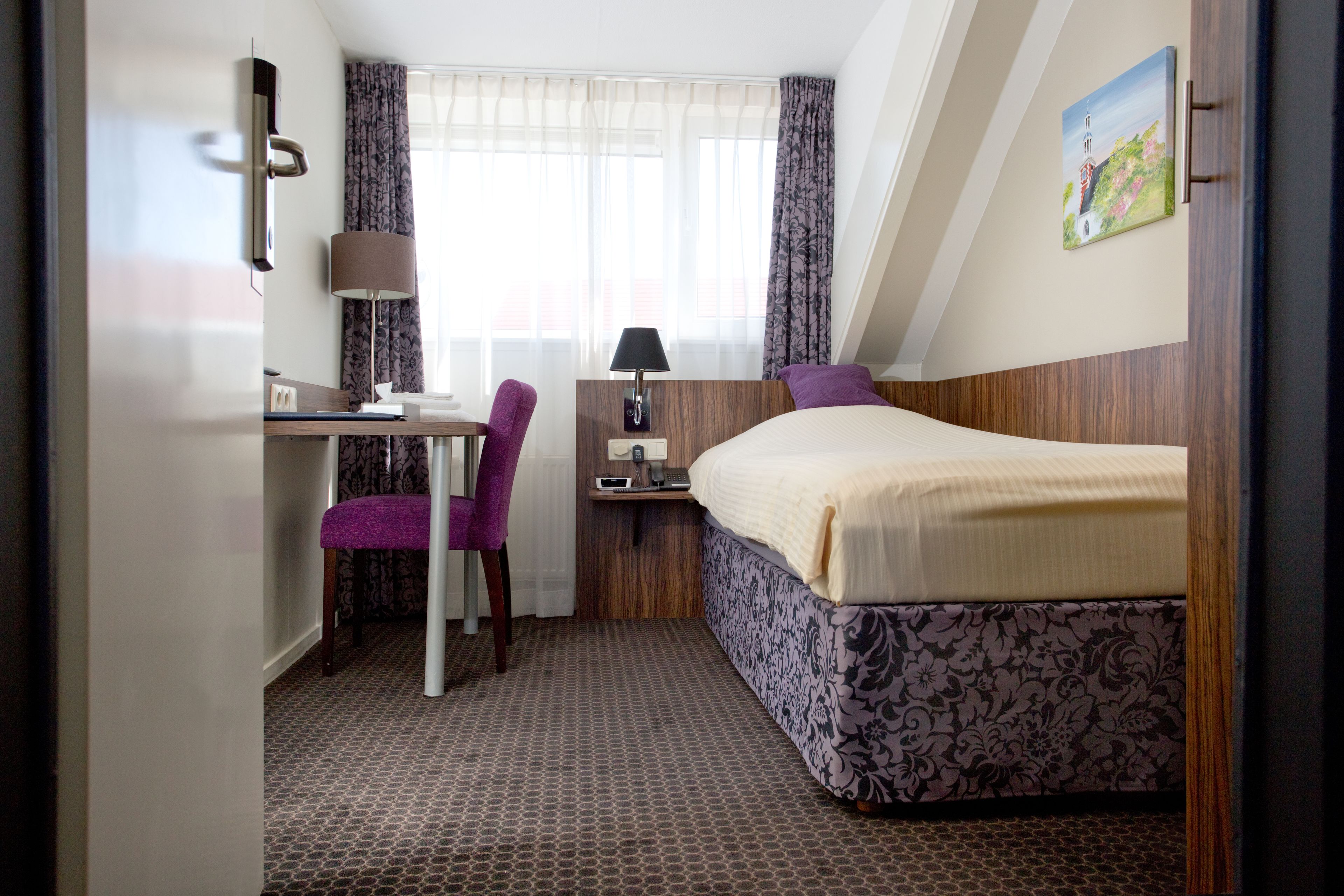 Quarto de hotel com cama, escrivaninha, cadeira, abajur e um quadro na parede.