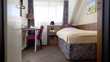 Quarto de hotel com cama, escrivaninha, cadeira, abajur e um quadro na parede.