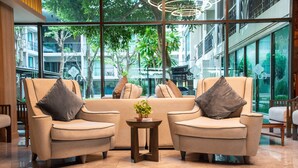 Um lobby moderno com duas poltronas bege, uma pequena mesa lateral de madeira com uma planta, e janelas amplas que oferecem vista dos edifícios e áreas verdes ao redor.