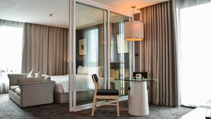 Quarto de hotel moderno com porta de vidro que dá para uma varanda, um sofá, uma cadeira, uma mesinha e um abajur.