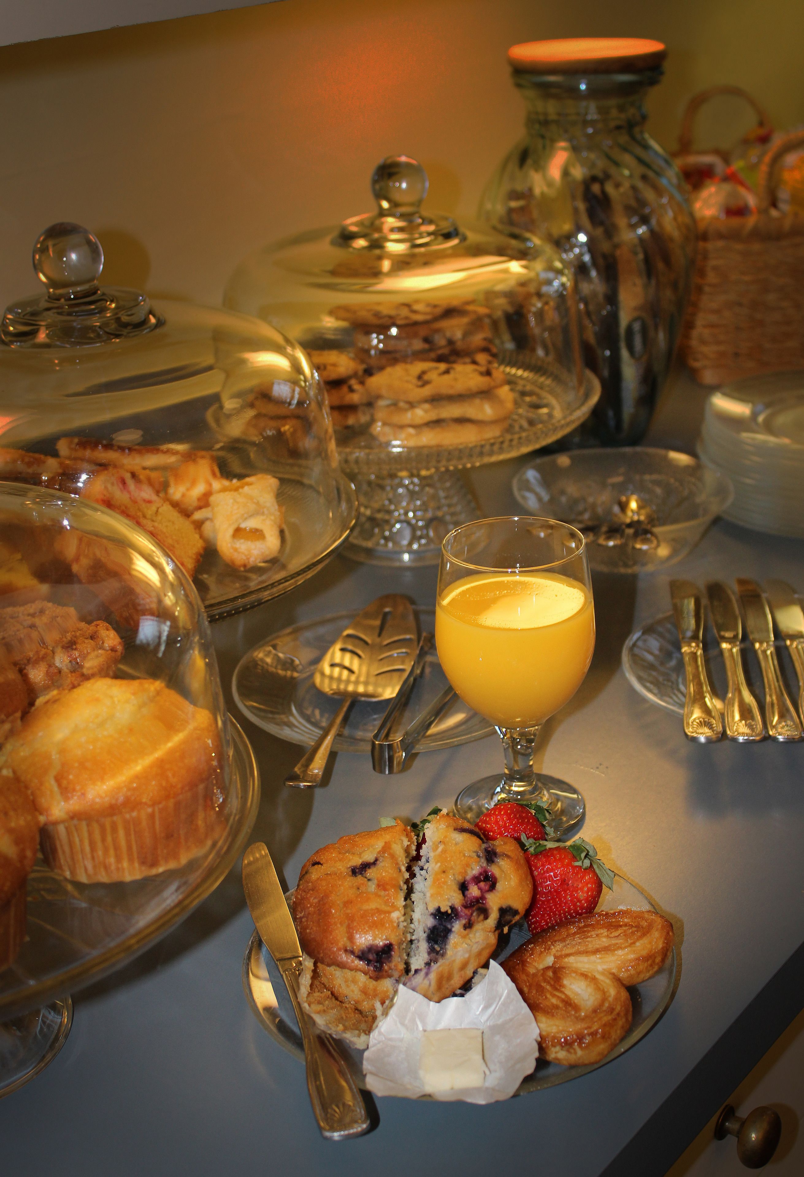 Um buffet de café da manhã com diversos pães e doces, um copo de suco de laranja e talheres.