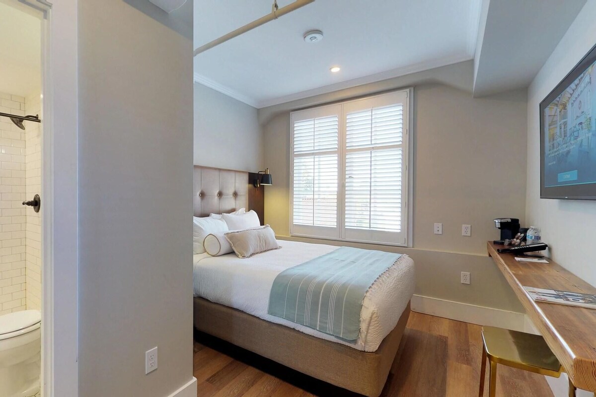 Quarto com cama, janela com persianas, escrivaninha com cadeira e área de chuveiro.