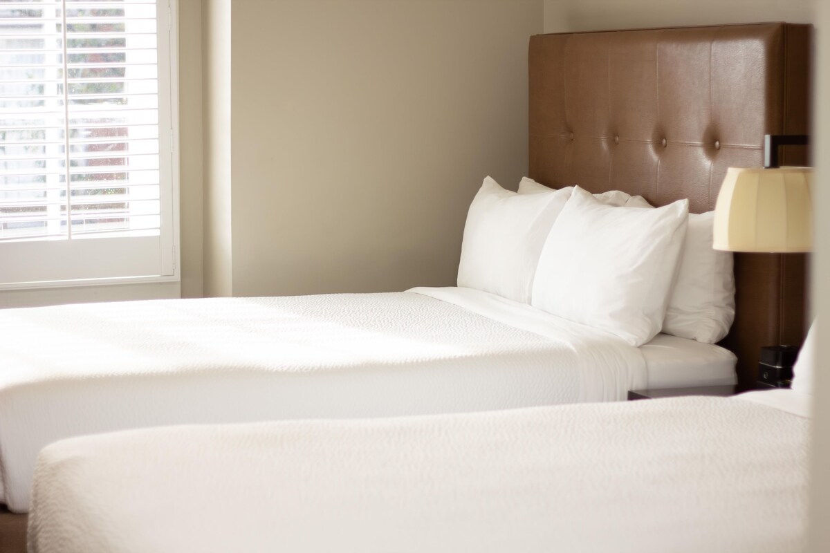 Quarto de hotel com uma cama grande, cabeceira e janela com persianas.