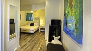 Quarto de hotel com cama, escrivaninha, televisão, um quadro e uma planta.