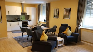 Sala de estar moderna com área de jantar, cozinha ao fundo, piso de madeira e grandes janelas com cortinas.
