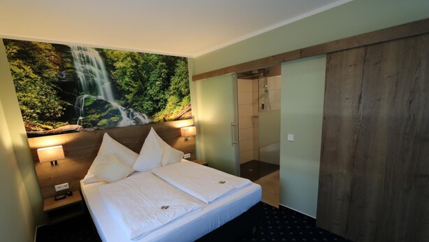 Quarto de hotel com cama, mesa de cabeceira, luminária na parede, área de chuveiro e um espelho grande.