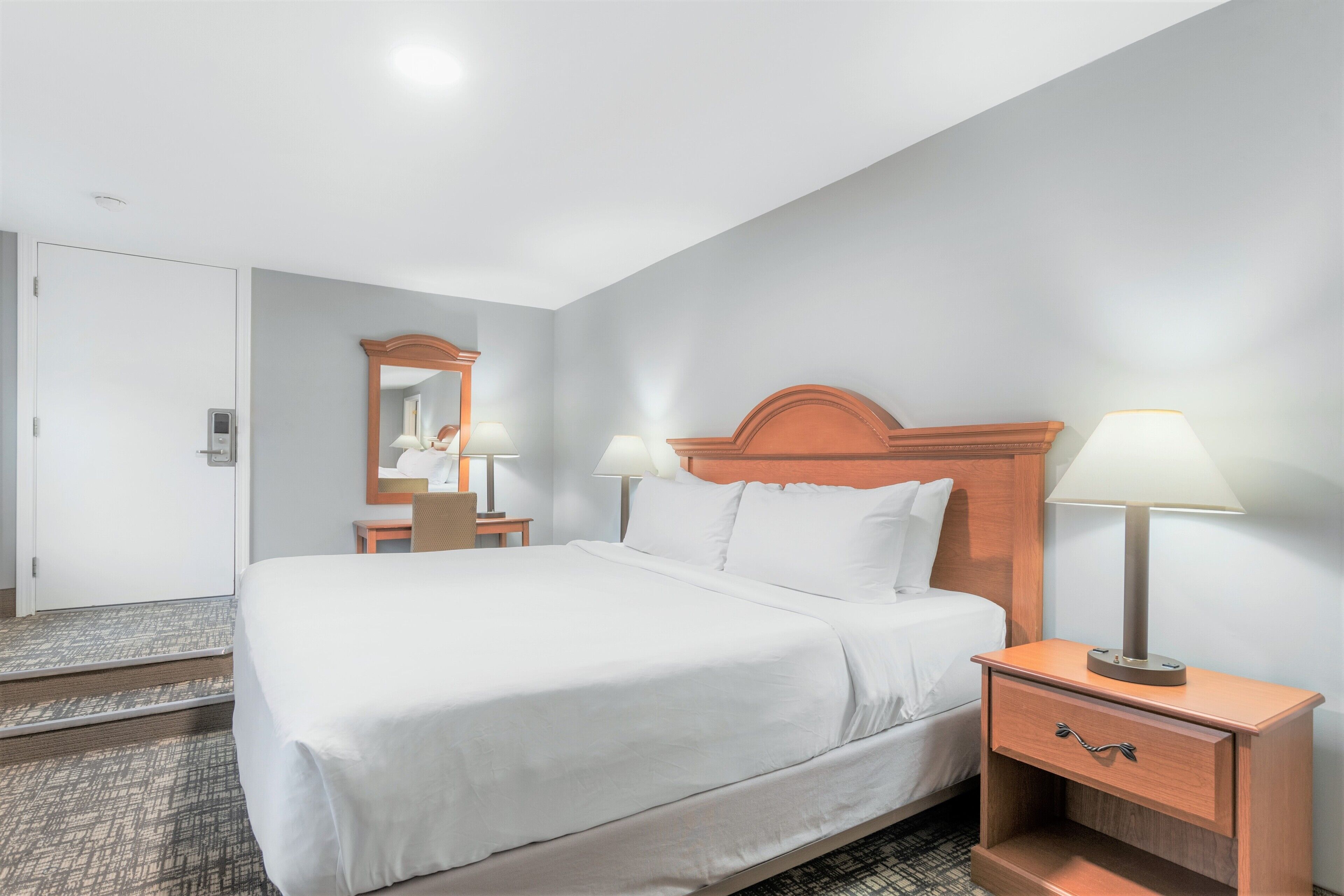 Quarto de hotel com cama, luminárias de cabeceira, mesa de apoio, espelho e porta.