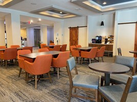 Um espaço de refeições de hotel com cadeiras e mesas laranja, uma estação de café e uma porta de madeira.