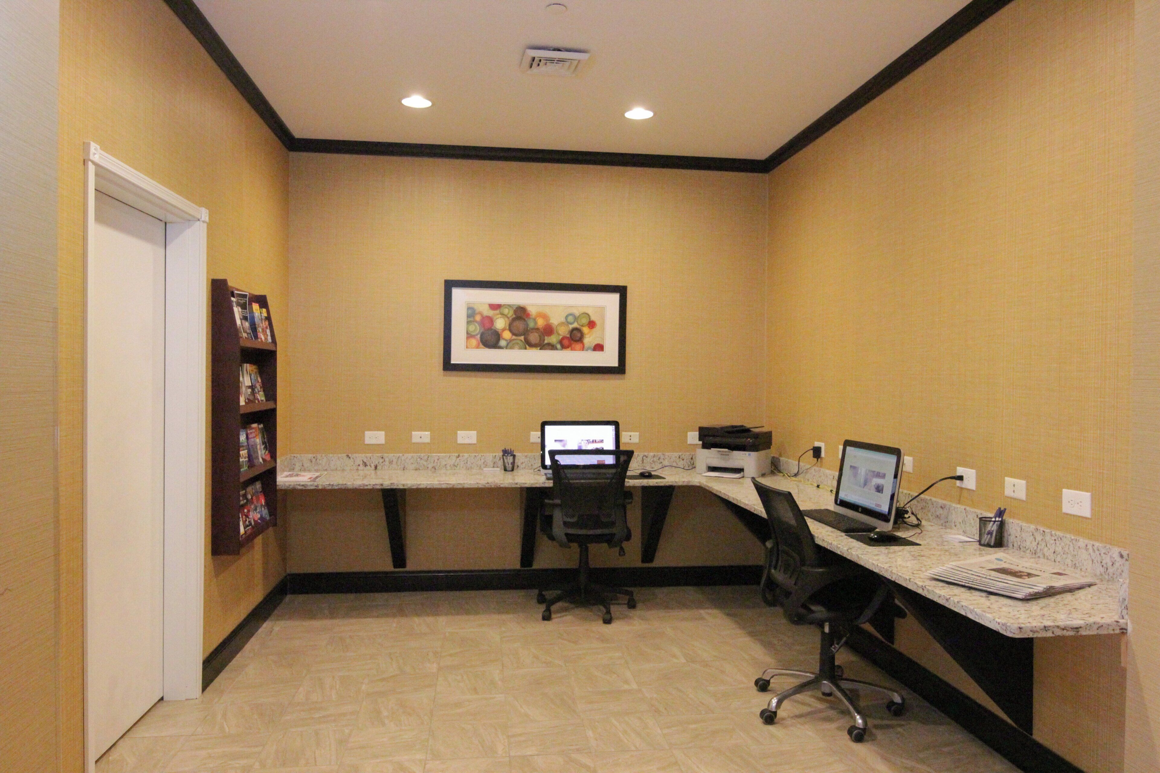 Quarto de hotel com uma escrivaninha comprida, dois computadores, uma cadeira e uma estante de livros.