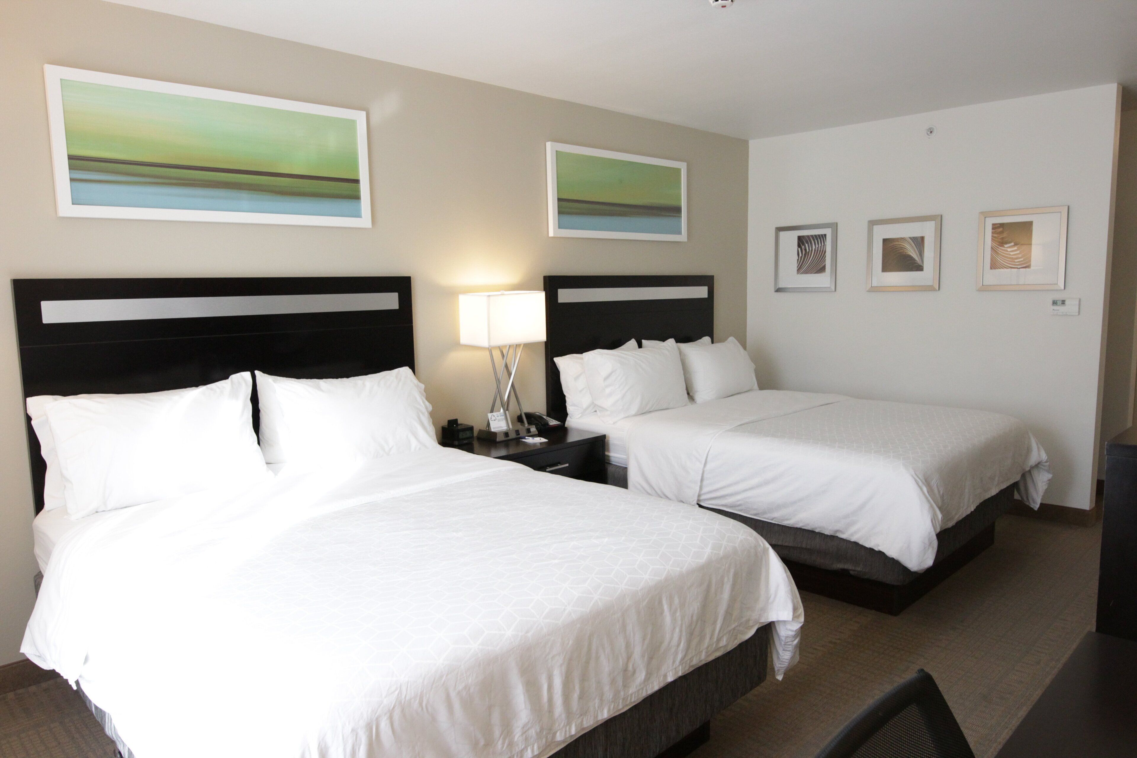 Quarto de hotel com duas camas, uma mesa de cabeceira, um abajur e três quadros na parede.