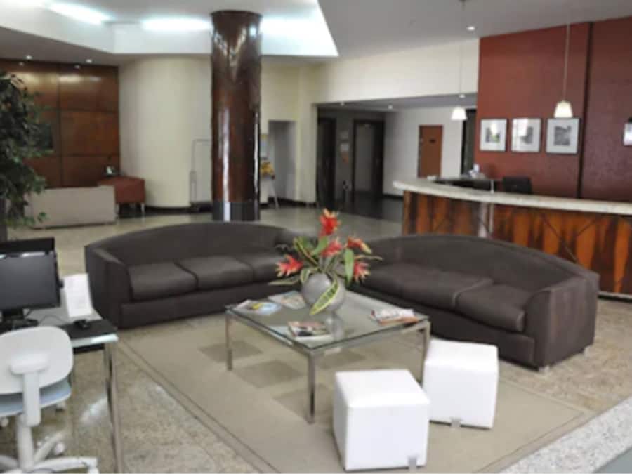 Um lobby moderno com um sofá marrom escuro curvo, uma mesa de centro de vidro com um vaso de flores, um balcão de recepção e uma cadeira de escritório branca.