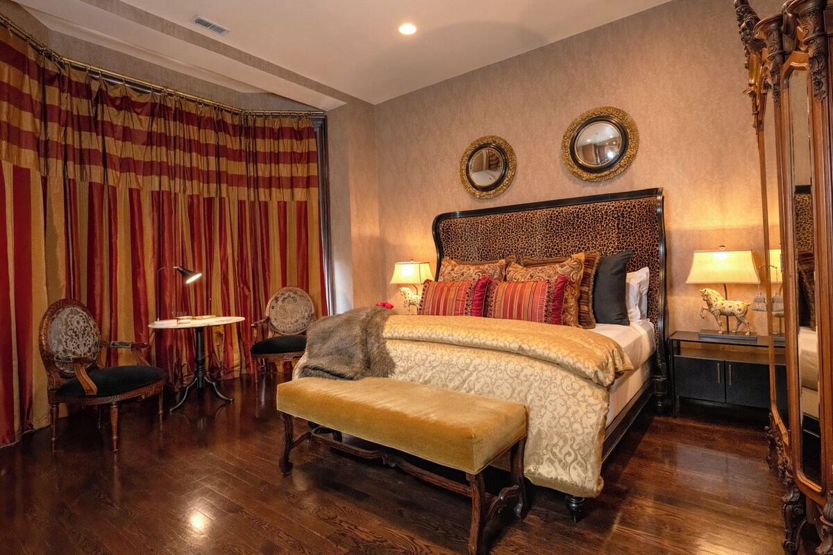 Quarto de hotel com uma cama grande, duas mesas de cabeceira, uma cadeira, uma mesa redonda pequena e um banco.