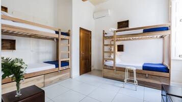 Quarto em dormitório com beliches, uma escrivaninha de madeira, um banquinho pequeno e um vaso de planta.