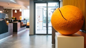 Um saguão moderno de hotel com uma mesa de recepção, uma grande escultura laranja e uma porta de vidro que dá para o exterior.