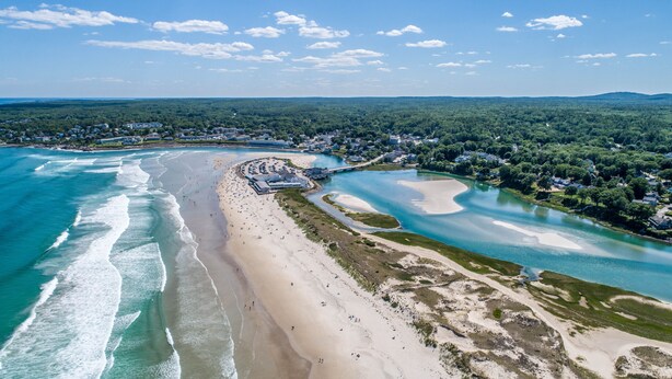 Vista aérea de uma área costeira com uma praia arenosa, um pequeno porto de iates e edifícios residenciais.