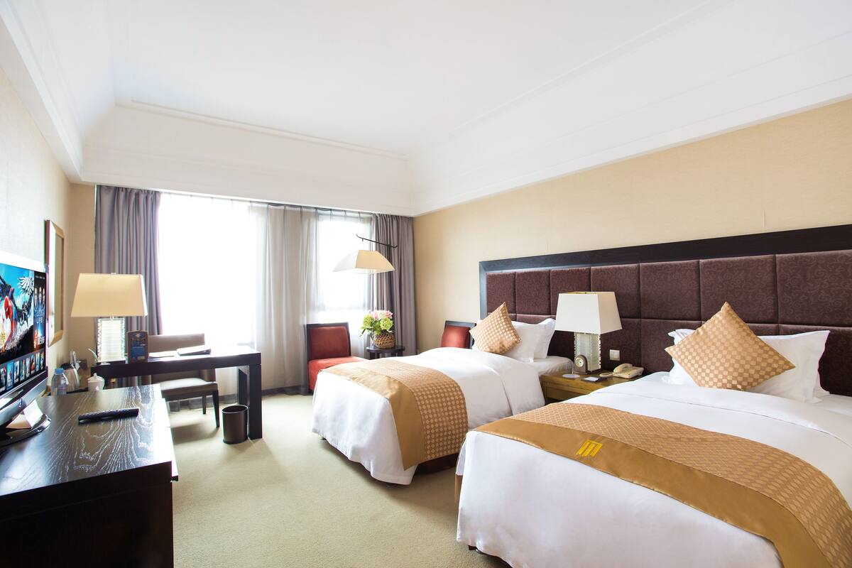 Quarto de hotel com duas camas, uma escrivaninha, uma televisão e uma janela com cortinas.
